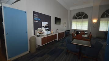 Um café moderno com um letreiro que diz “Preciso. De. Café.” e uma área de estar com mesas e cadeiras.