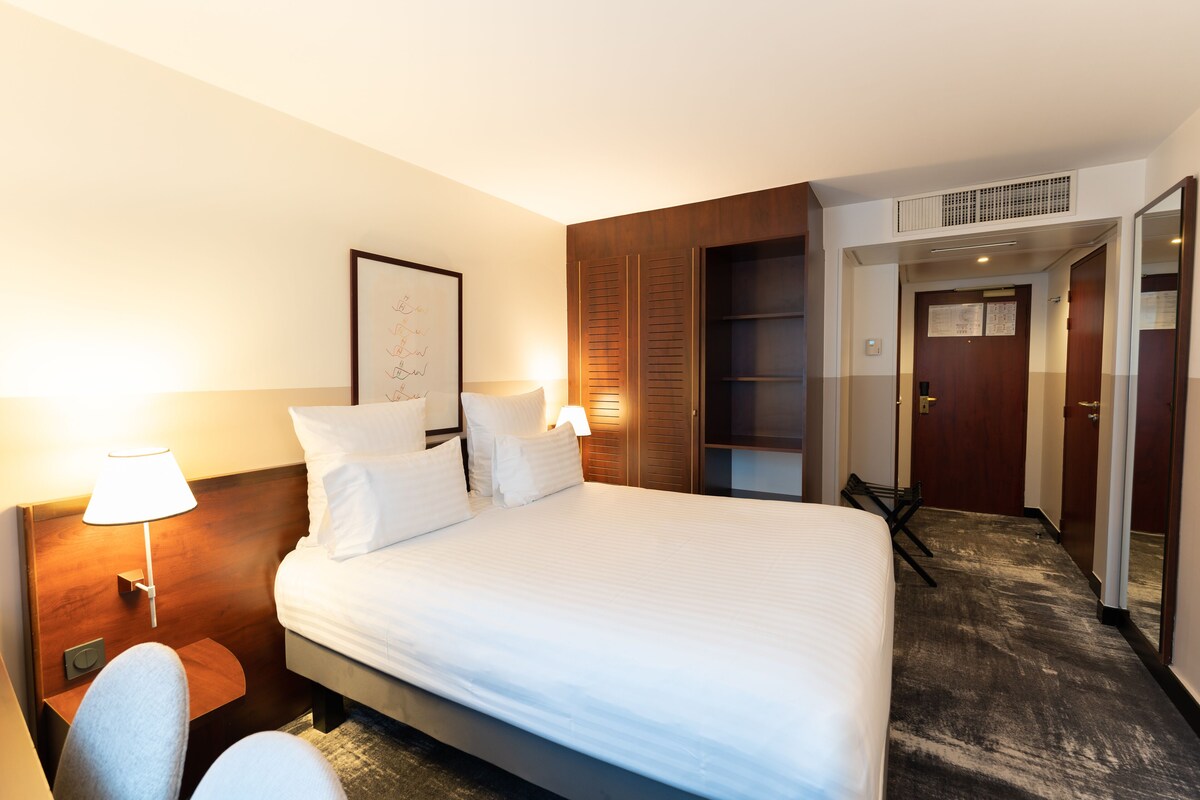 Quarto de hotel com uma cama grande, abajures, cabeceira, uma obra de arte emoldurada, guarda-roupa de madeira e piso coberto por carpete.