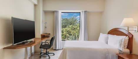 Quarto de hotel com televisão de tela plana sobre uma escrivaninha de madeira, uma cadeira de escritório, duas camas com roupa de cama branca, uma mesa de cabeceira com abajur e uma janela com cortinas.