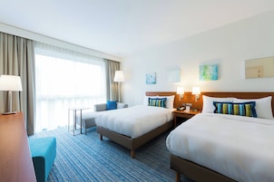 Quarto de hotel com duas camas, uma escrivaninha e uma cadeira. Há duas mesas de cabeceira com luminárias. O quarto possui uma janela grande com cortinas.