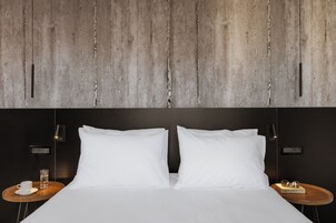 Quarto de hotel moderno com cabeceira de concreto, roupa de cama branca e duas mesas de cabeceira com luminárias e um copo de água.