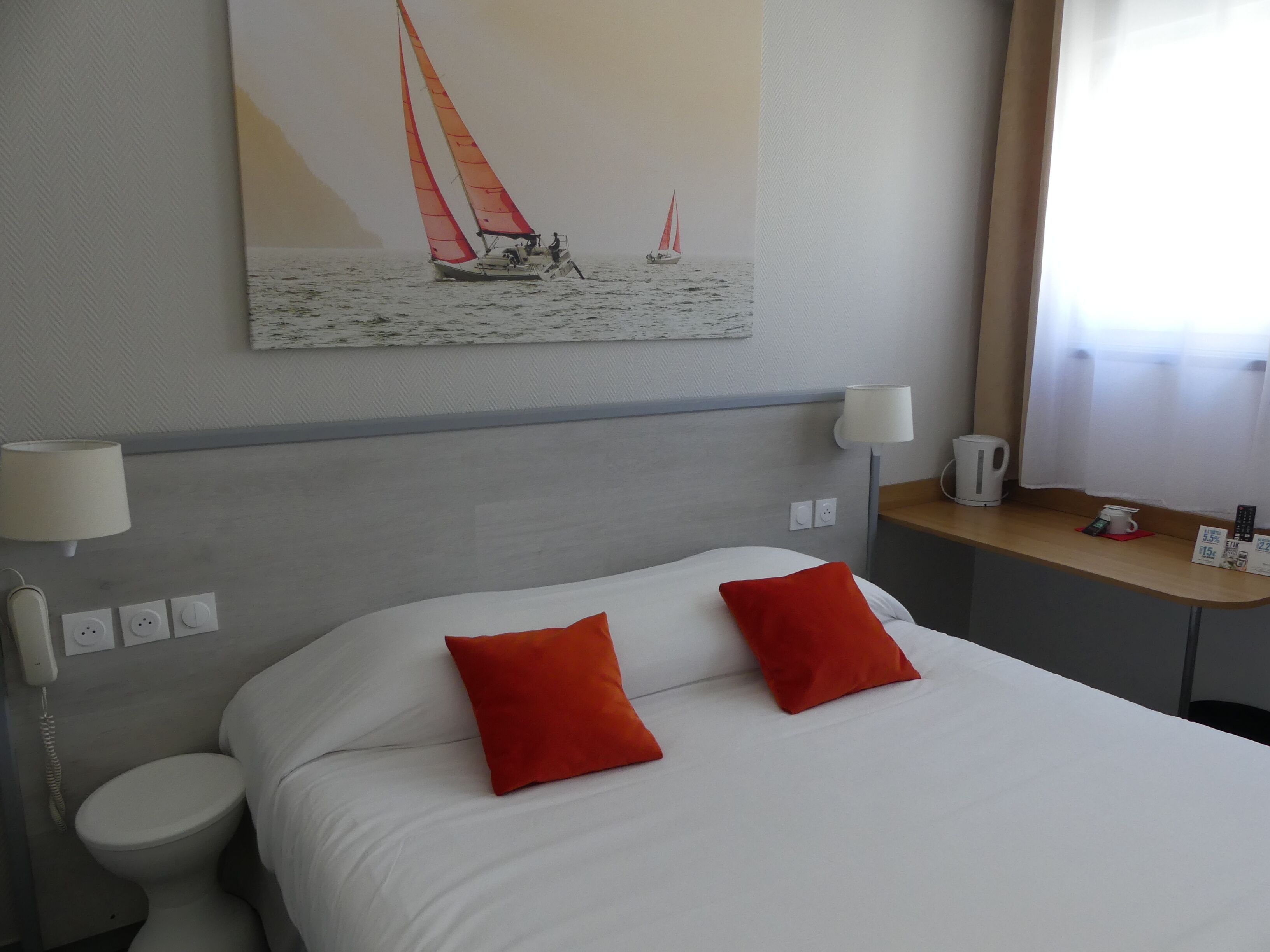 Quarto de hotel com cama, duas almofadas vermelhas, pintura de veleiros na parede, abajures, mesa de cabeceira com luminária, chaleira e xícara.