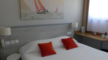 Quarto de hotel com cama, duas almofadas vermelhas, pintura de veleiros na parede, abajures, mesa de cabeceira com luminária, chaleira e xícara.