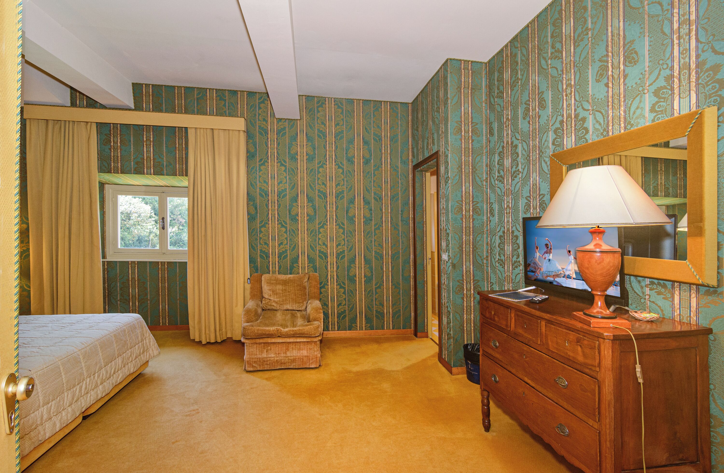 Quarto com cama, uma cadeira, cômoda, televisão e janela com cortinas.
