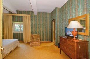Quarto com cama, uma cadeira, cômoda, televisão e janela com cortinas.