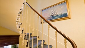 Uma escadaria com balaústres brancos e corrimão de madeira, que leva a um patamar com um quadro emoldurado de um navio a vela.