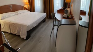 Quarto de hotel com cama, escrivaninha, cadeira, abajur e cortinas.