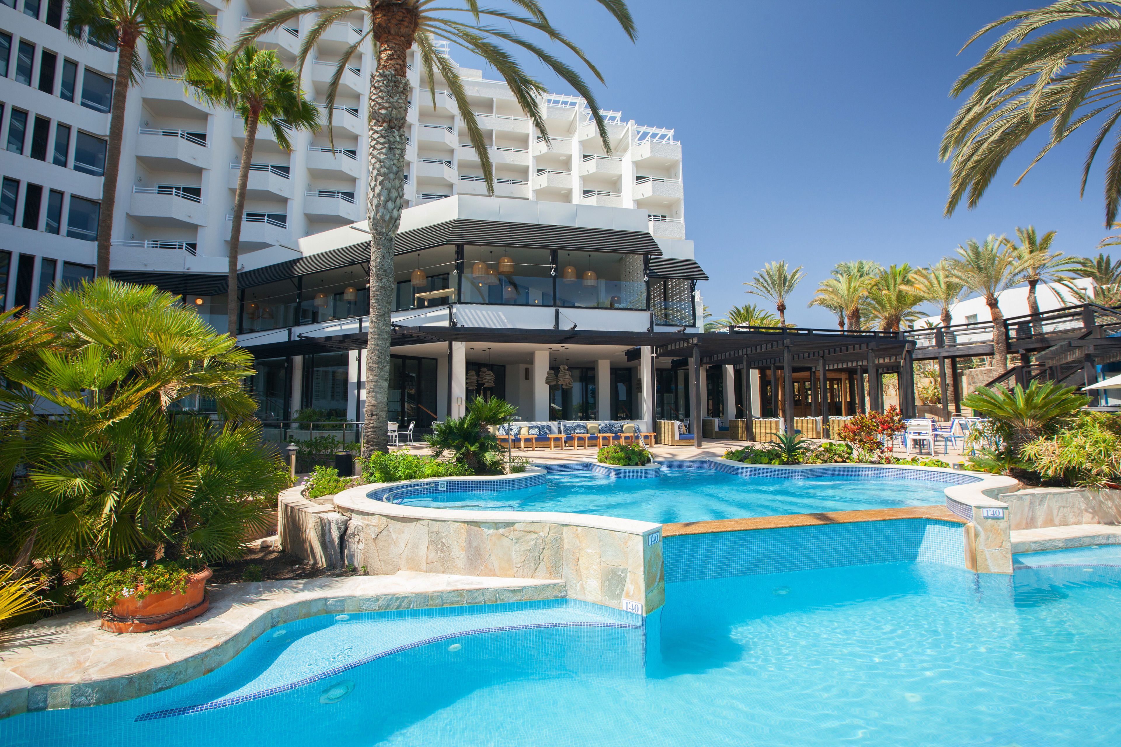 Um hotel com área de piscina, palmeiras e um edifício moderno com varandas.
