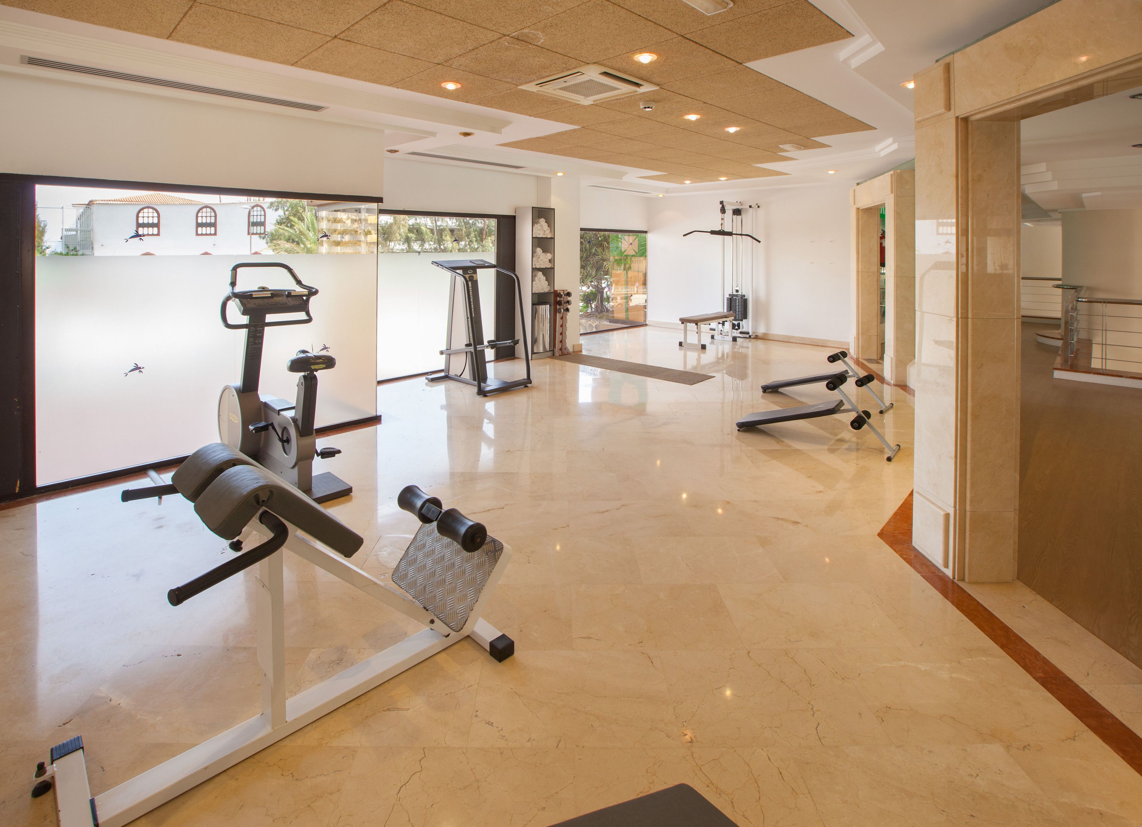 Uma academia espaçosa com diversos equipamentos de ginástica, um espelho grande e vista para um edifício lá fora.