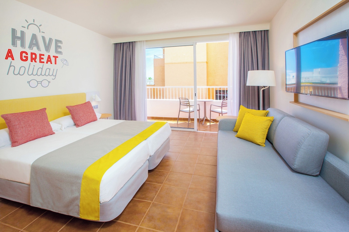 Quarto de hotel com cama, sofá, varanda com mesa e cadeiras, e parede com mensagem de férias.