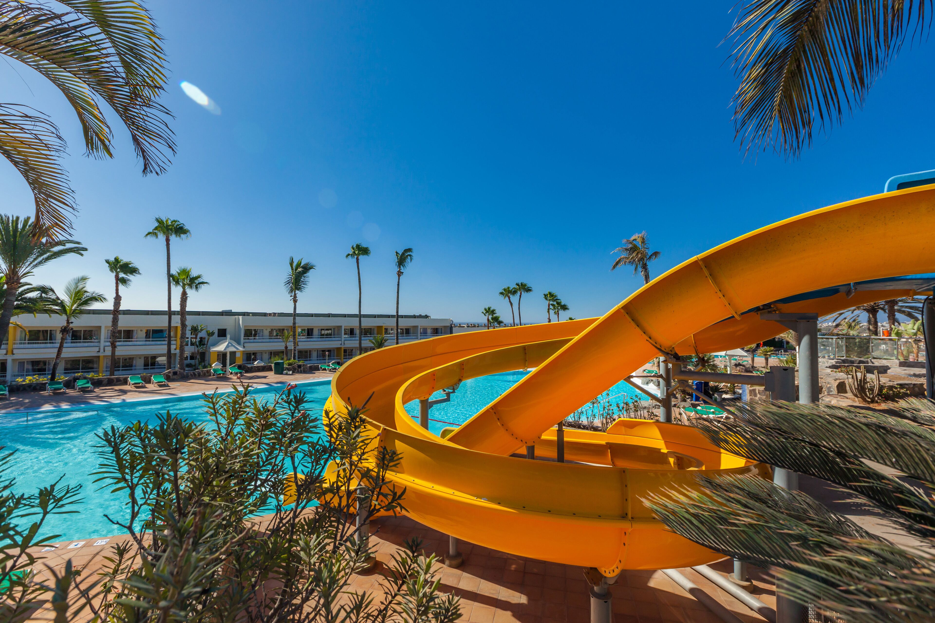 Área de piscina com um escorregador amarelo, palmeiras e um céu azul límpido.