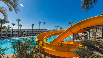 Área de piscina com um escorregador amarelo, palmeiras e um céu azul límpido.
