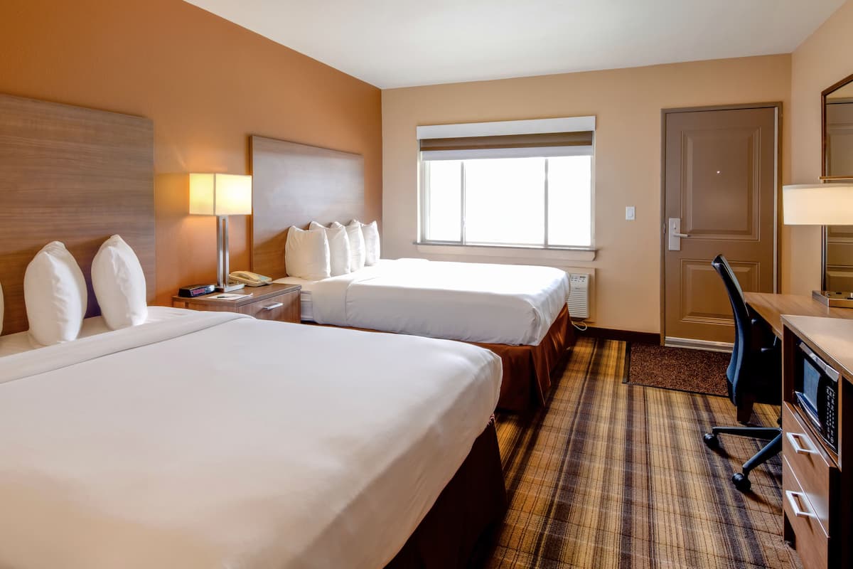 Quarto de hotel com duas camas, uma escrivaninha, uma cadeira, um abajur e uma janela.