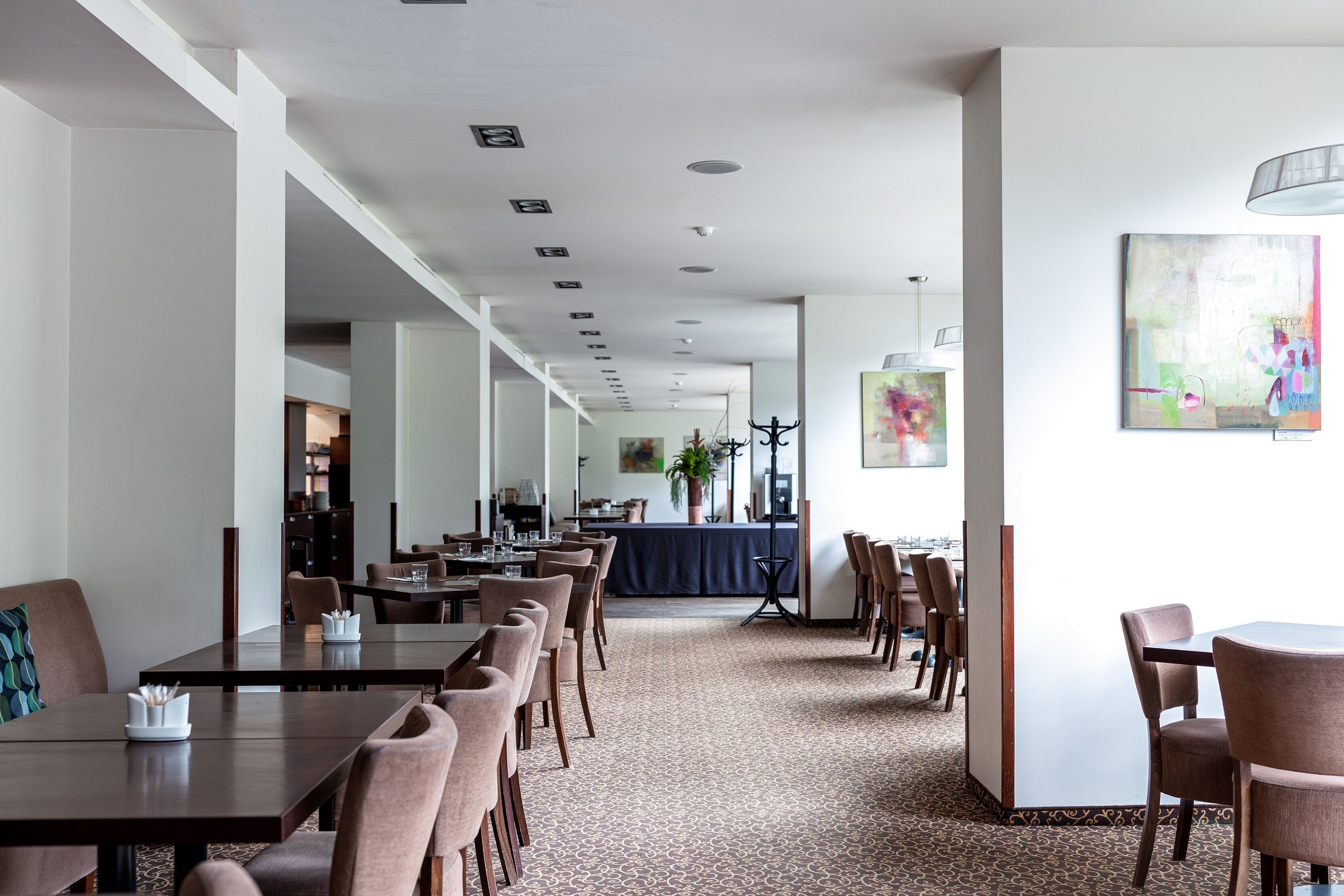 Uma sala de jantar de hotel com mesas e cadeiras compridas, um bar e obras de arte nas paredes.