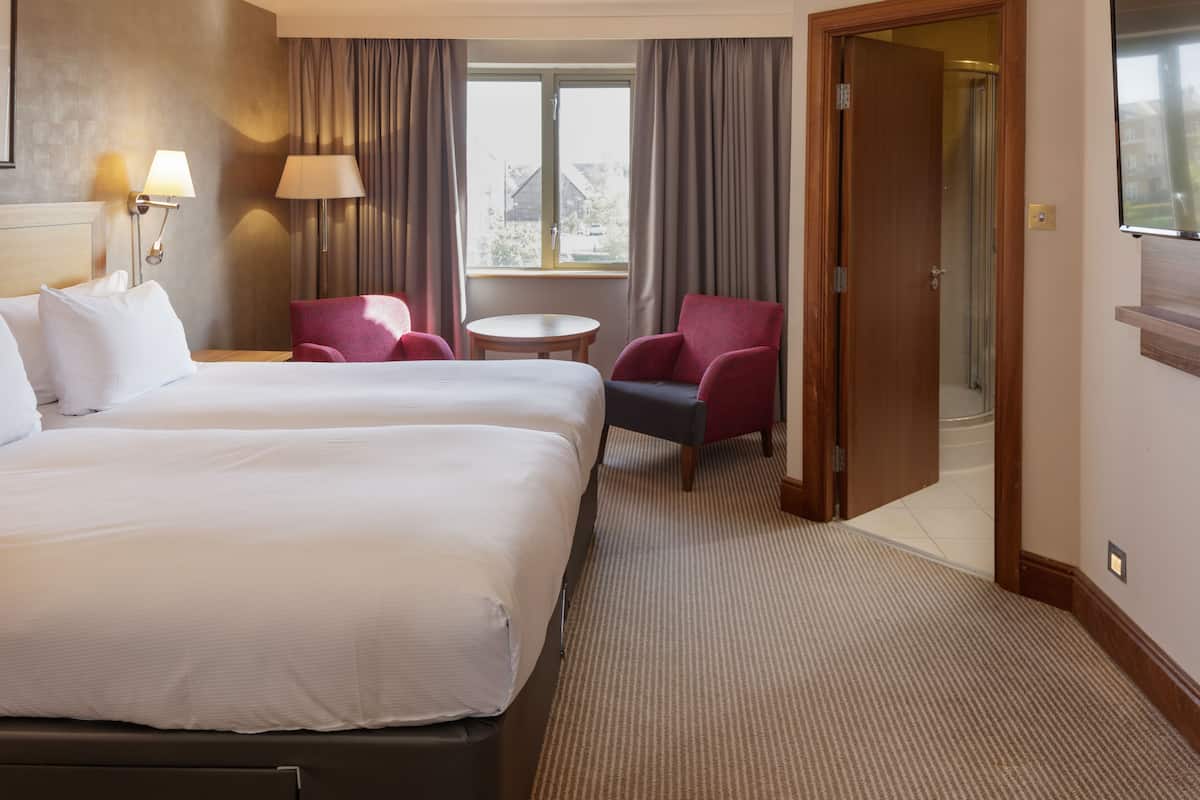 Quarto de hotel com duas camas, um espaço de estar com mesa e cadeiras, uma janela com cortinas e área de chuveiro.