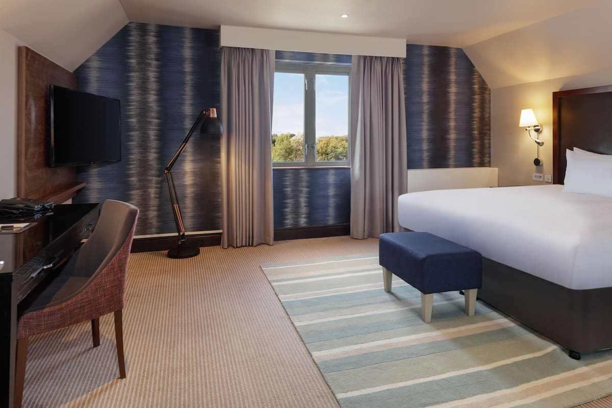Quarto de hotel com cama, escrivaninha, cadeira, televisão e janela com cortinas.