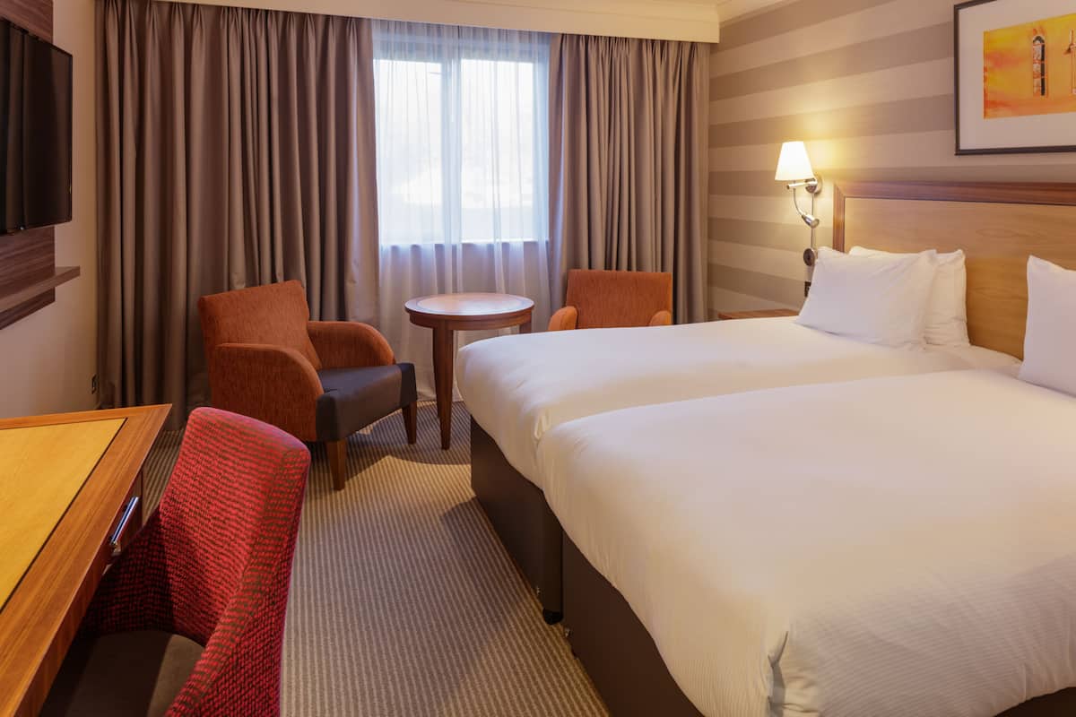 Quarto de hotel com cama, escrivaninha, duas poltronas, mesinha, televisão e abajur.