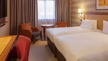 Quarto de hotel com cama, escrivaninha, duas poltronas, mesinha, televisão e abajur.