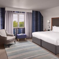 Quarto de hotel com cama, cadeira, pequena mesa, abajur e vista para área verde lá fora.