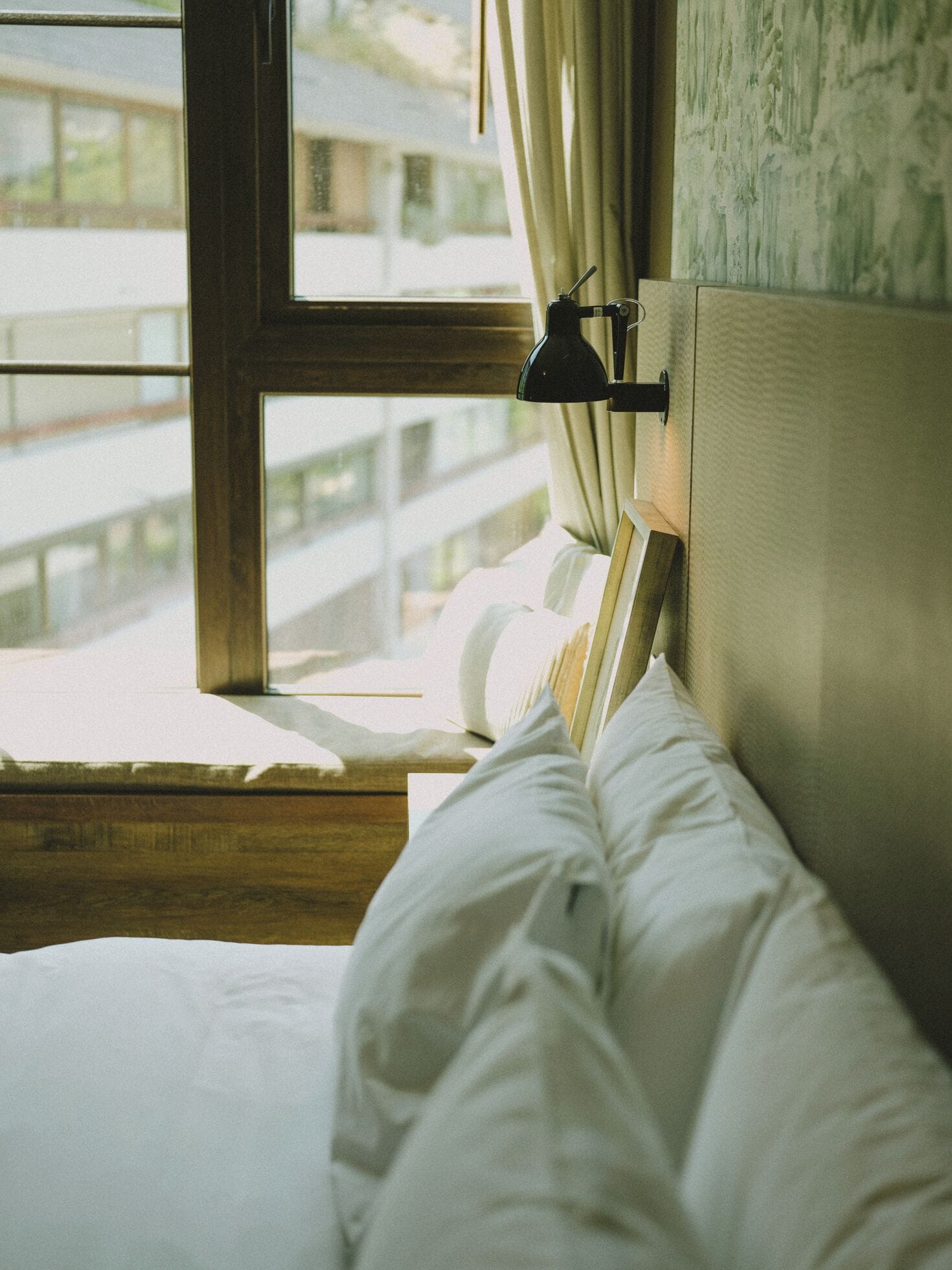 Uma cama com roupa de cama branca, uma luminária de mesa preta e uma janela com cortinas.