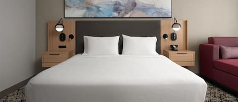 Quarto de hotel com cama grande, duas mesas de cabeceira com luminárias, uma poltrona vermelha e um quadro abstrato pendurado na parede.