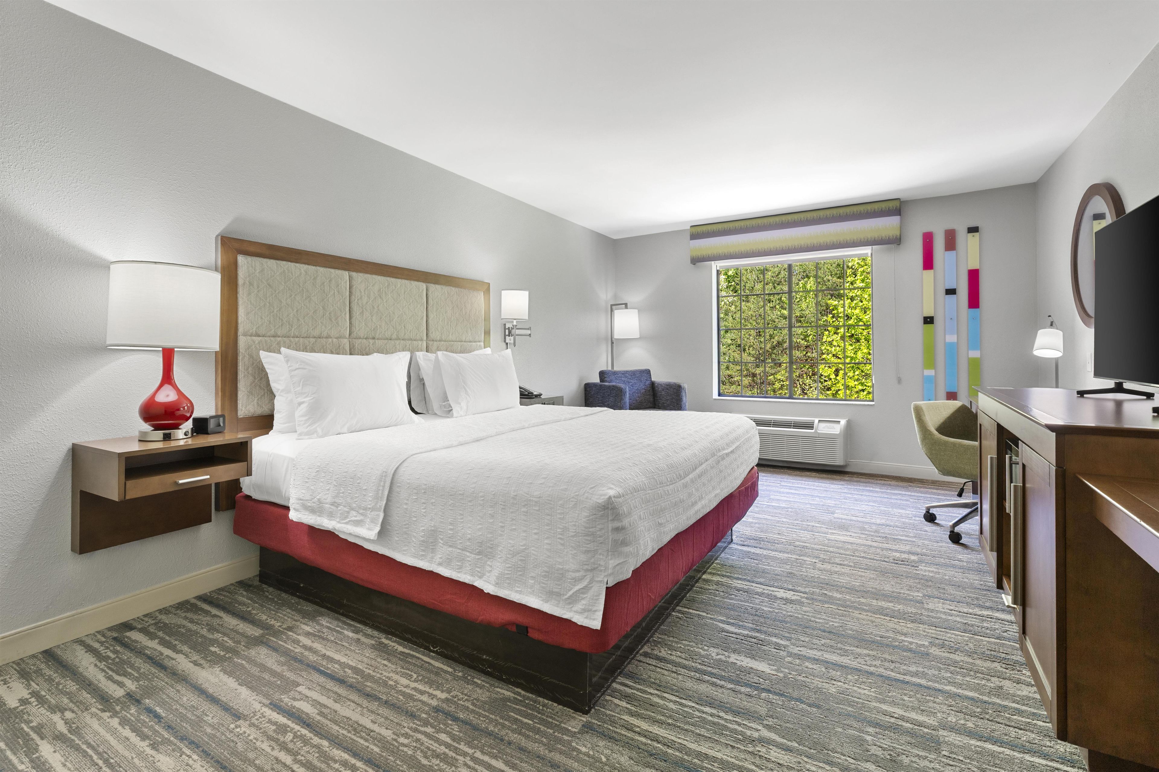 Quarto de hotel com cama, mesa de cabeceira, abajur, escrivaninha, cadeira e uma janela grande.