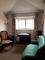 Quarto com sofá verde, cadeira de madeira, escrivaninha com papéis, janela com cortinas e radiador.