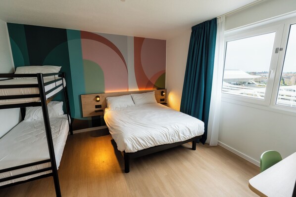 Quarto de hotel com beliche, uma cama de solteiro, mesa de cabeceira, abajur, janela com cortinas e vista para a cidade.