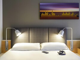 Quarto de hotel com cama, duas luminárias de cabeceira, um smartphone e um par de fones de ouvido na cama.