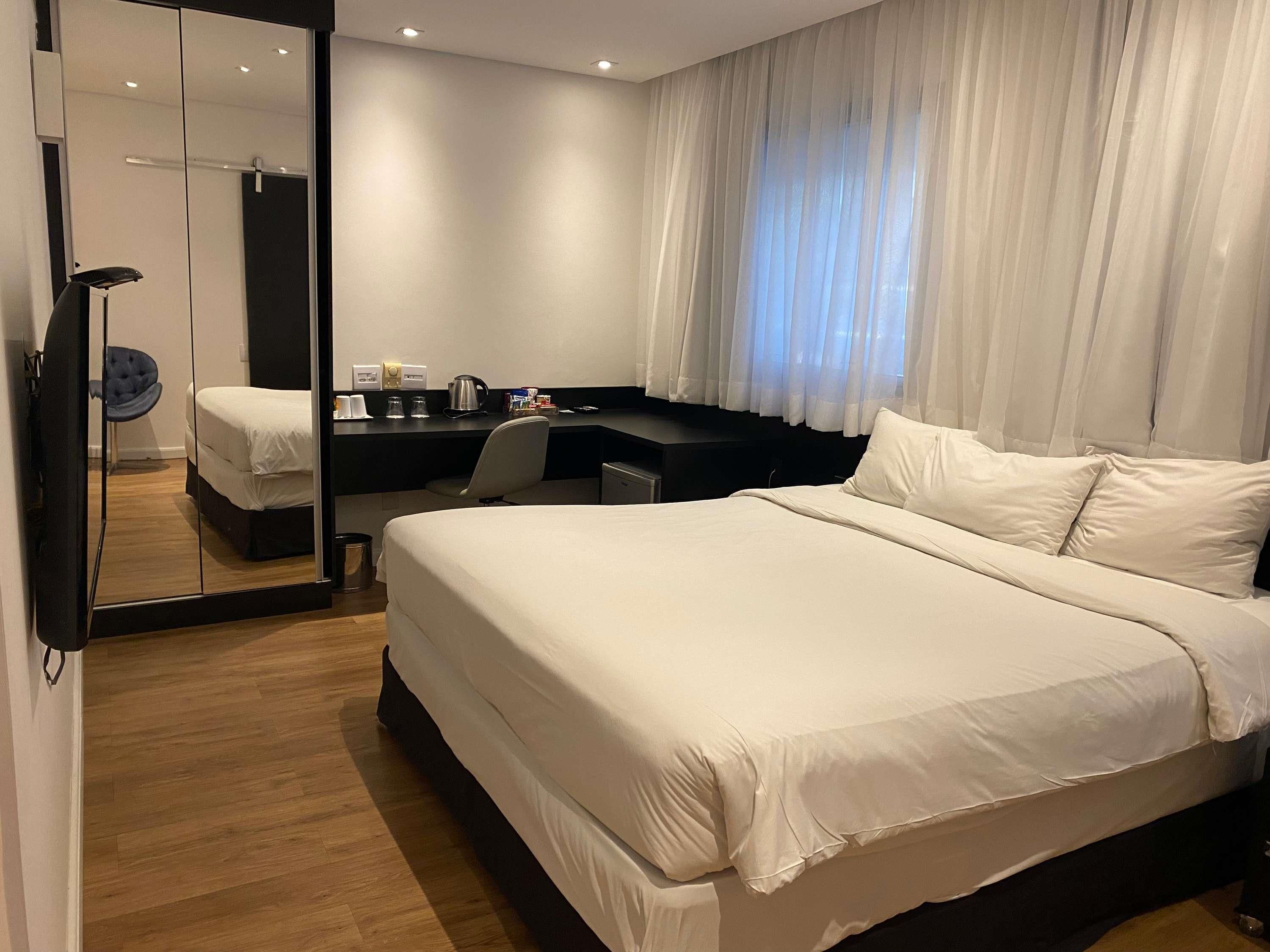 Quarto de hotel moderno com uma cama grande, uma escrivaninha, uma cadeira, um espelho e uma janela com cortinas leves.
