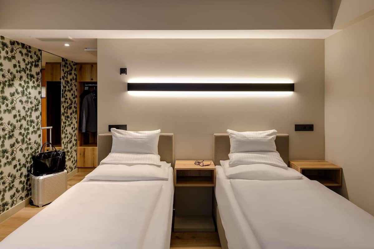 Quarto de hotel com duas camas de solteiro, mesa de cabeceira com copos, um cabideiro e um guarda-roupa com papel de parede floral.