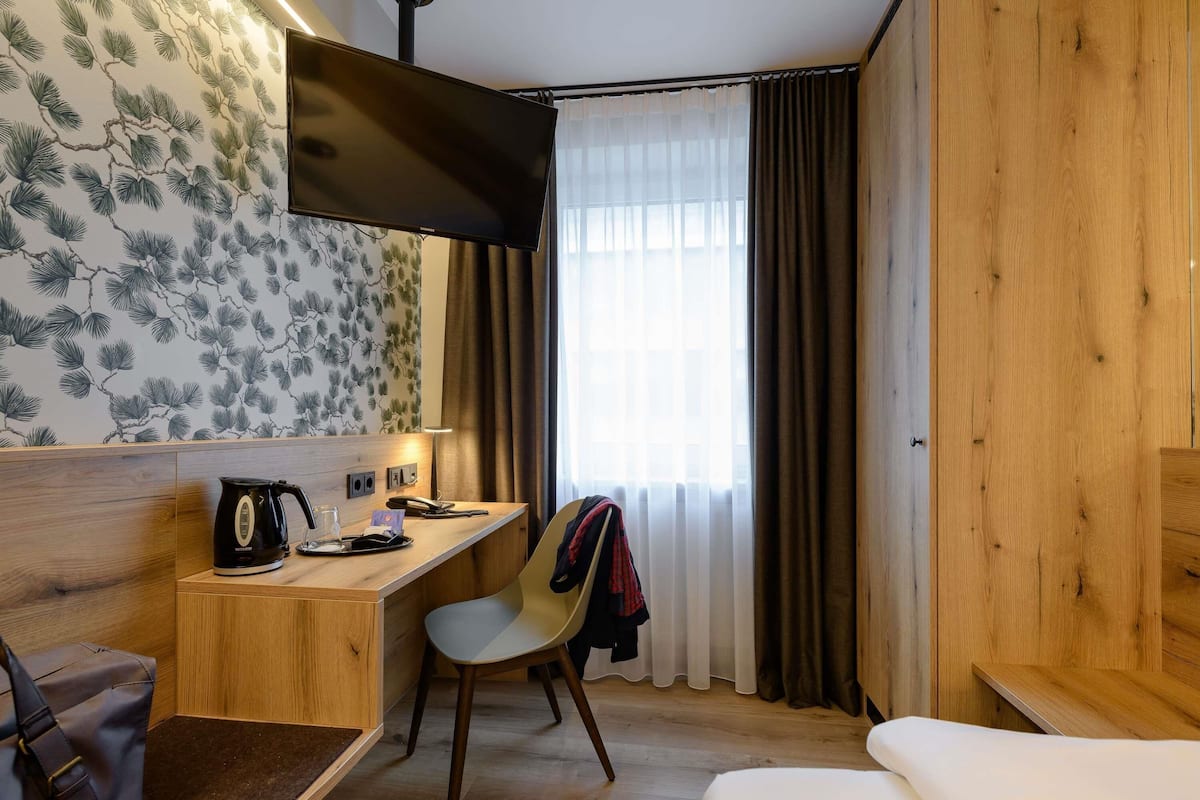 Quarto de hotel com escrivaninha de madeira, uma cadeira, uma cama, janela com cortinas e parede com padrão de folhas.