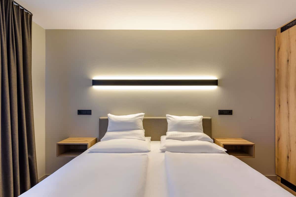 Cama de casal com lençóis brancos, duas mesas de cabeceira, uma luminária na parede e um guarda-roupa de madeira.