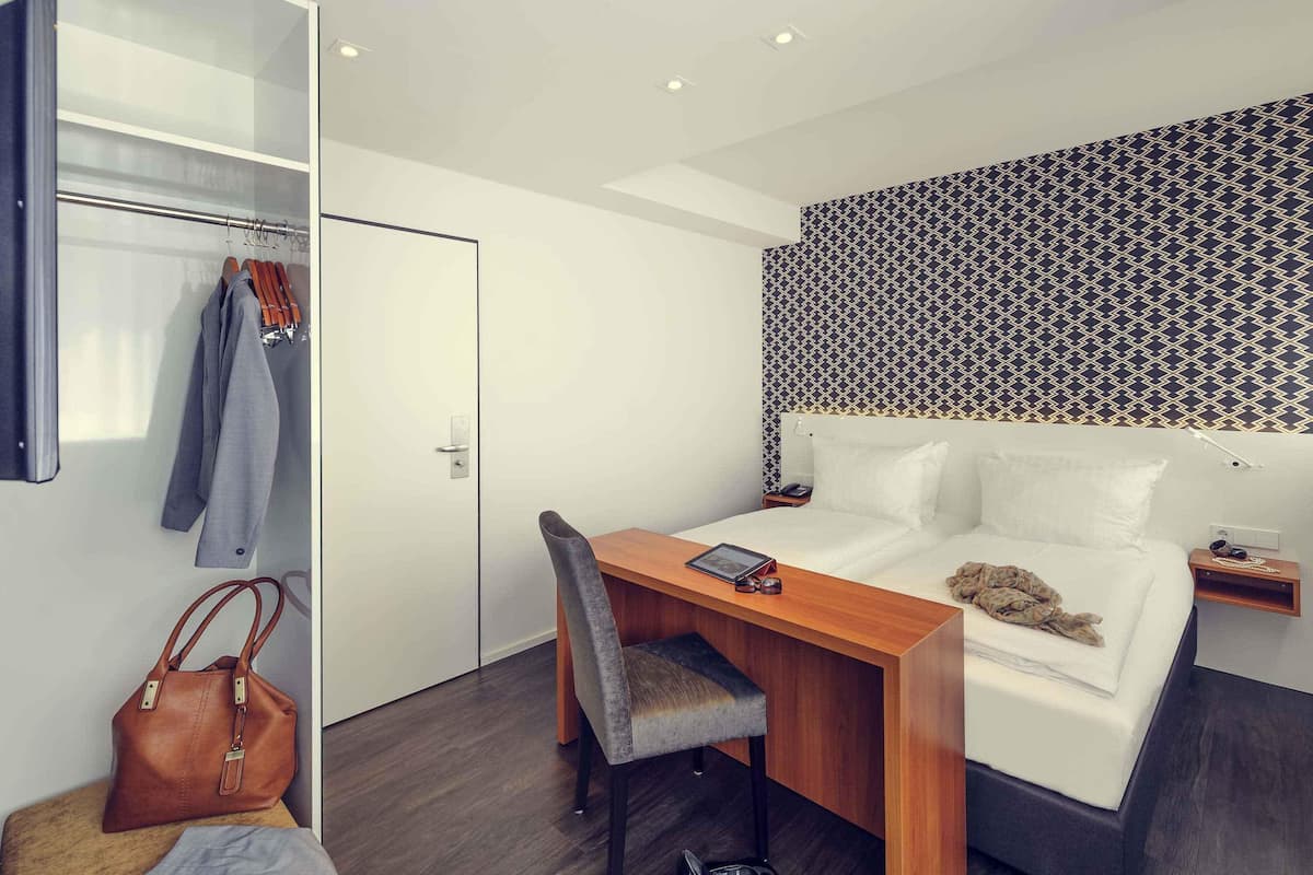 Quarto de hotel com cama, escrivaninha, cadeira, guarda-roupa e uma bolsa.