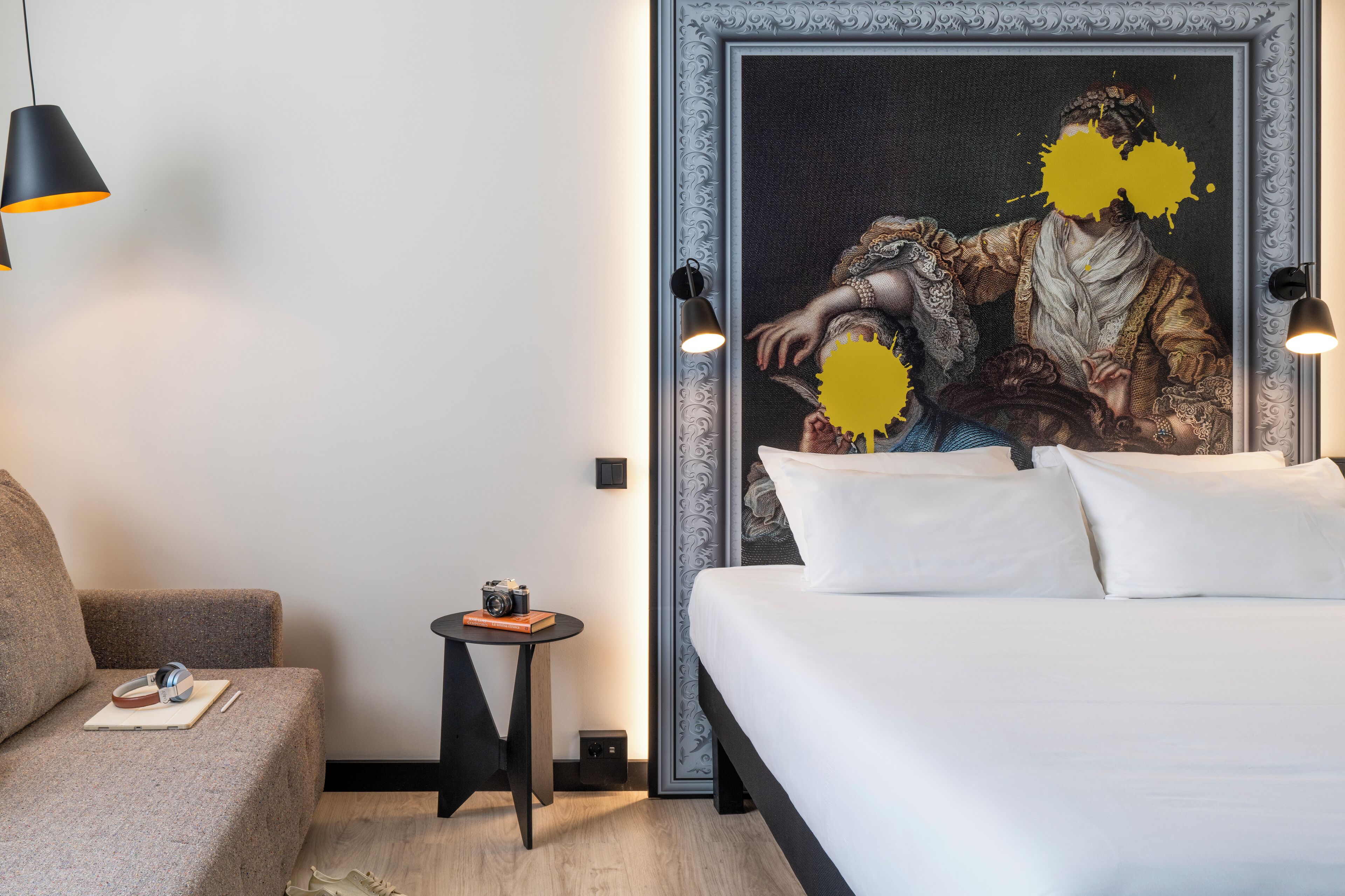 Quarto de hotel com uma cama, uma mesa pequena com uma câmera e um quadro pendurado na parede.