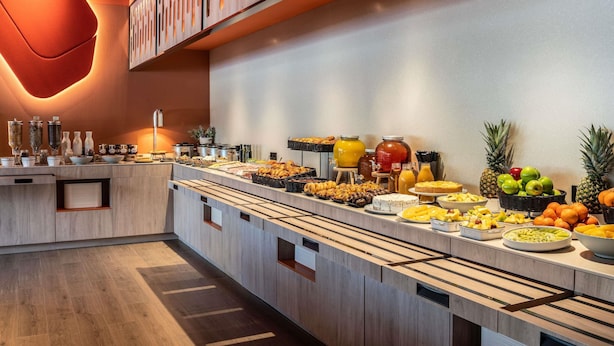 Buffet com diversas opções de comida e bebidas, incluindo frutas e doces, em ambiente moderno com painéis de madeira e luminária que emite luz quente.