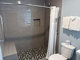 Área de chuveiro com divisória de vidro, cortina branca e vaso sanitário com rolo de papel higiênico.