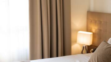 Quarto de hotel com cama bem arrumada, luminária em uma mesa de cabeceira e janela com cortinas.