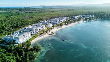 Vista aérea de um resort costeiro com vilas na orla marítima, águas cristalinas e colinas verdejantes ao fundo.
