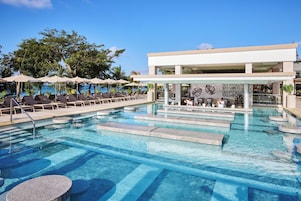 Piscina com espreguiçadeiras e guarda-sóis, área de bar e um edifício com design moderno.