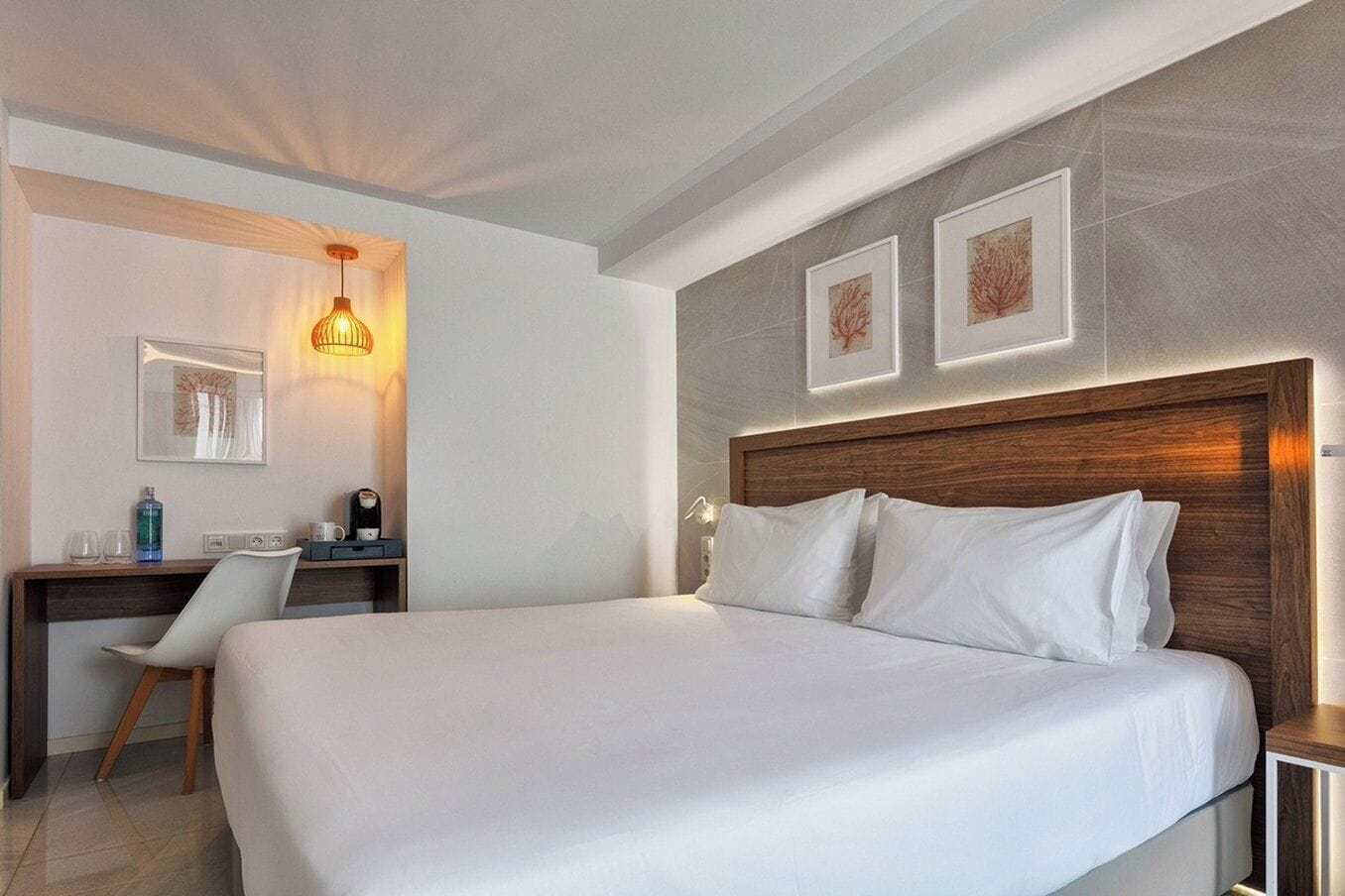 Quarto de hotel com uma cama grande, uma escrivaninha com cadeira, mesa de cabeceira e uma luminária na parede.