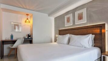 Quarto de hotel com uma cama grande, uma escrivaninha com cadeira, mesa de cabeceira e uma luminária na parede.