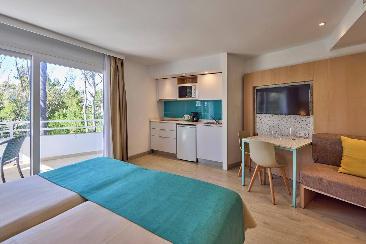 Quarto de hotel com cama, escrivaninha, sofá, cozinha compacta e varanda com uma cadeira.