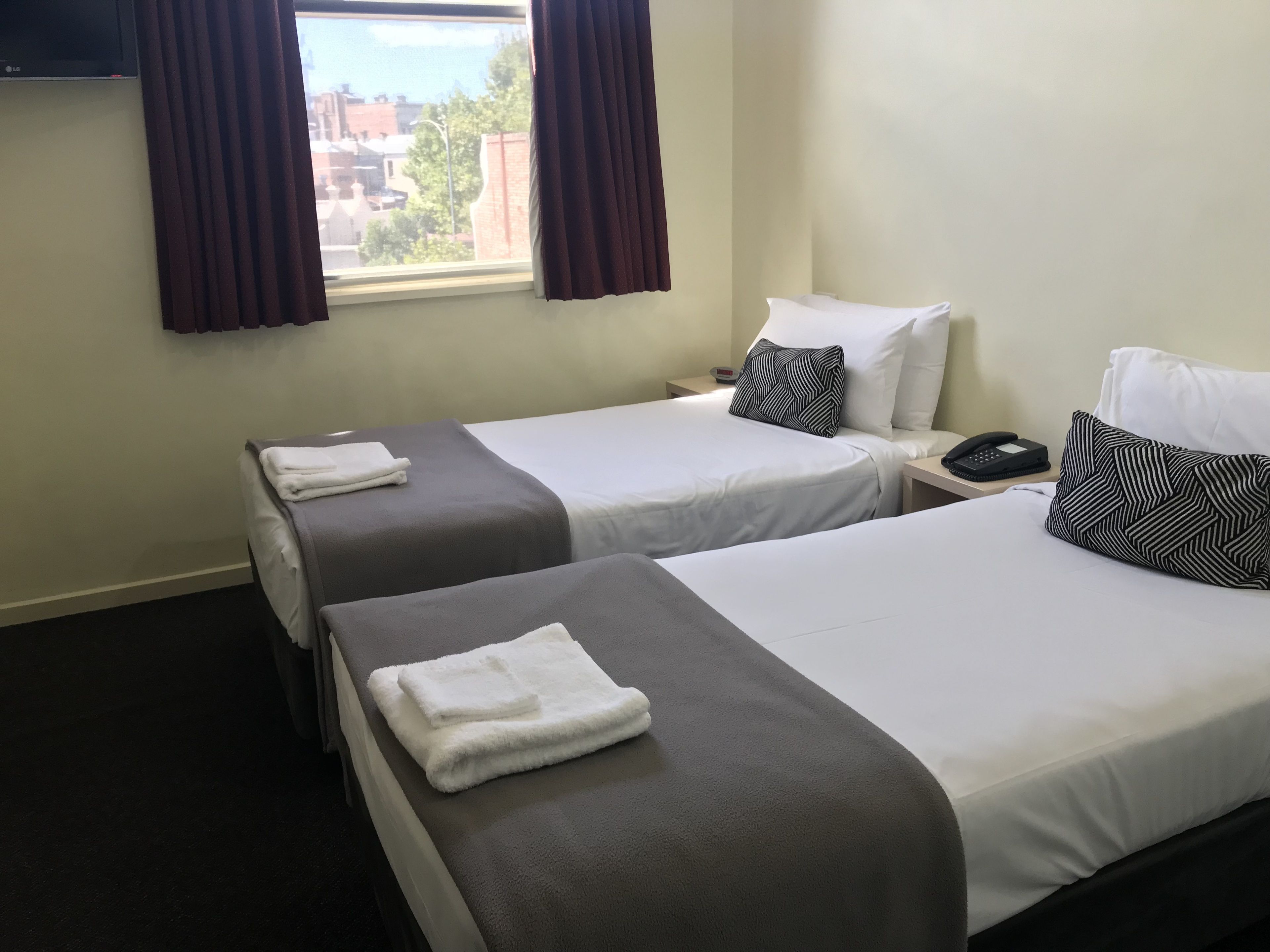 Quarto de hotel com duas camas, cada uma com edredom branco e manta cinza. Há duas almofadas, uma com padrão preto e branco. É visível uma janela com cortinas, e uma televisão de tela plana está montada na parede.