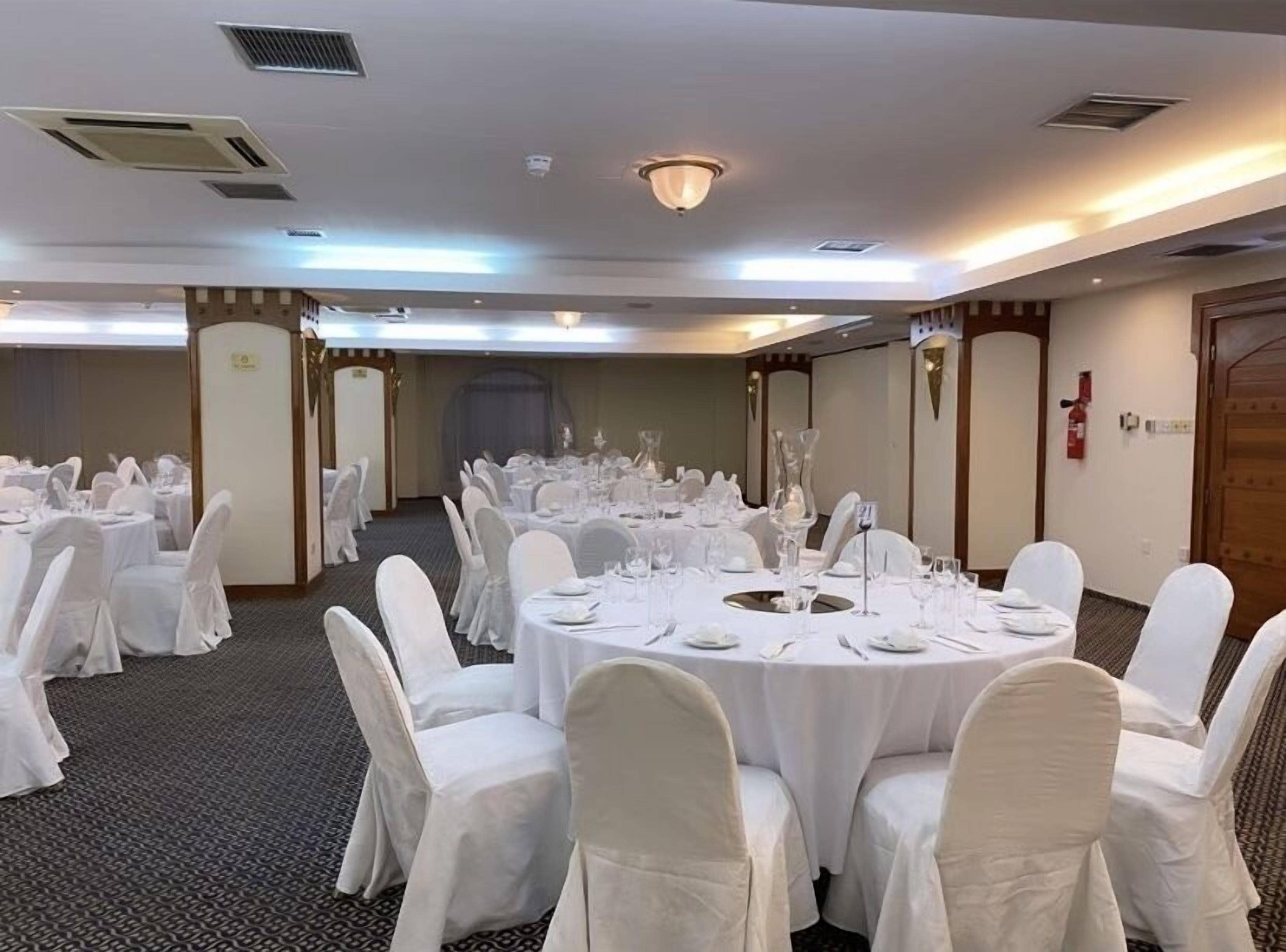 Uma sala de banquetes com mesas redondas preparadas para refeições, cadeiras brancas e um extintor de incêndio na parede.