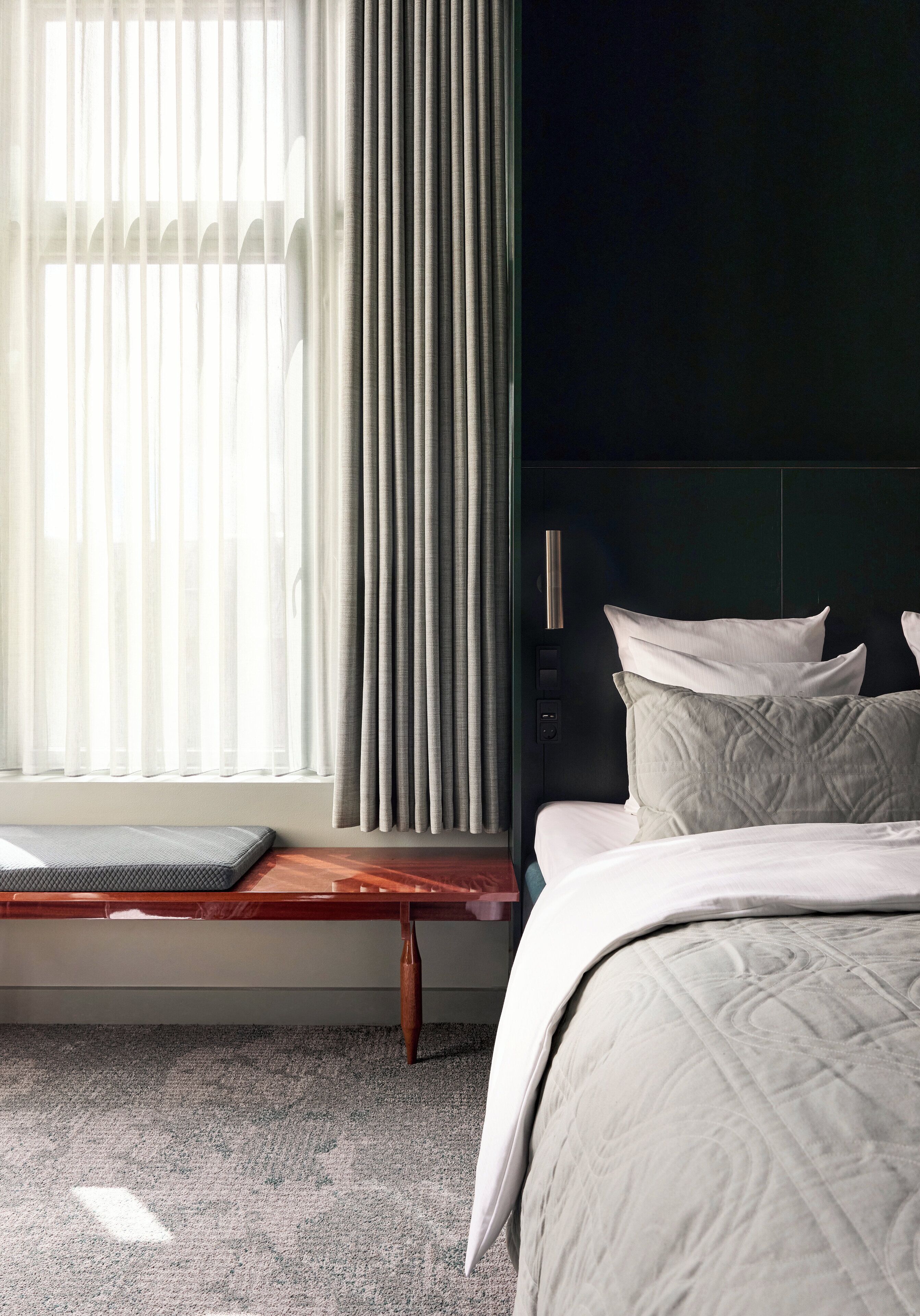 Quarto de hotel com cama, um banco junto à janela e cabeceira escura.