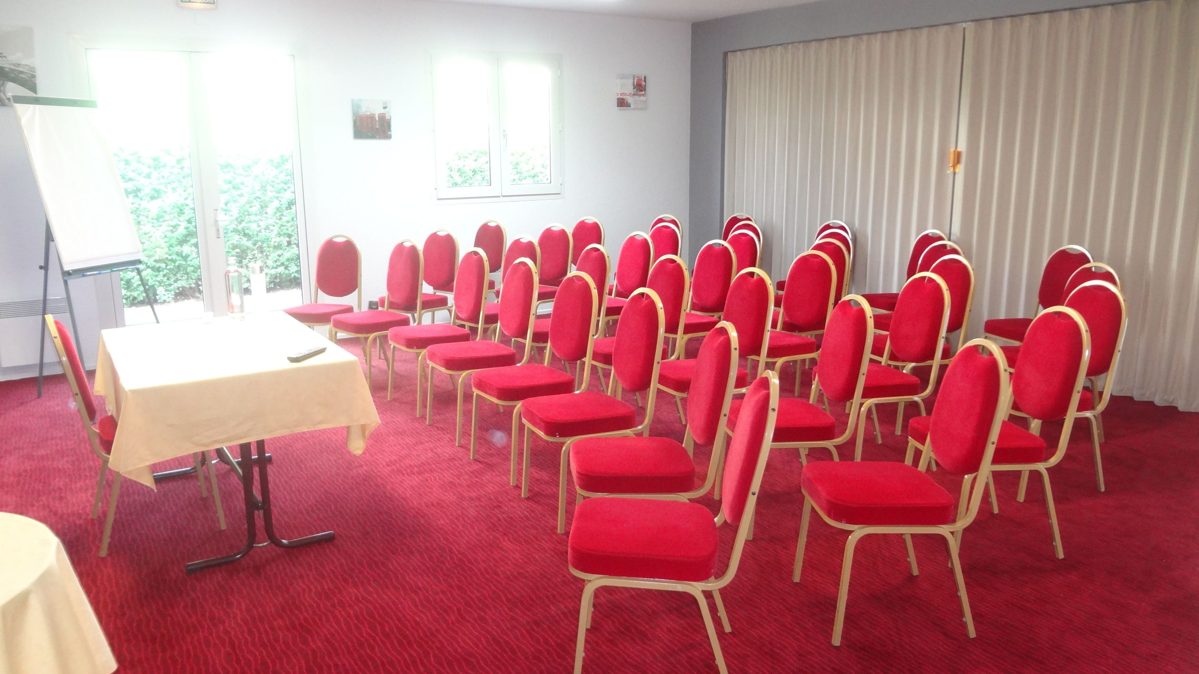 Uma sala de conferências com cadeiras vermelhas dispostas em fileiras, uma mesa com toalha branca e janelas amplas com cortinas.