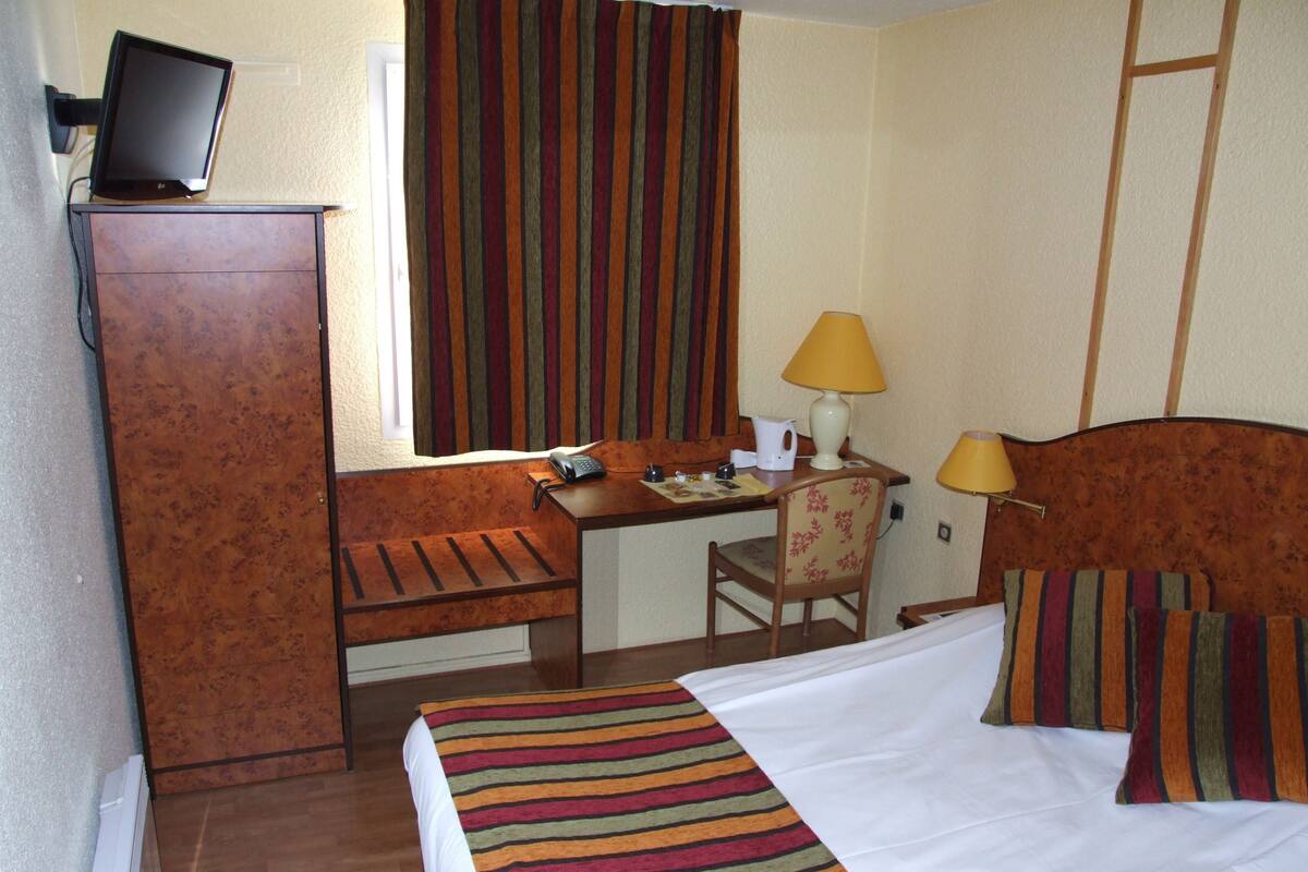 Quarto de hotel com uma cama, escrivaninha, cadeira, luminária e televisão sobre um suporte.