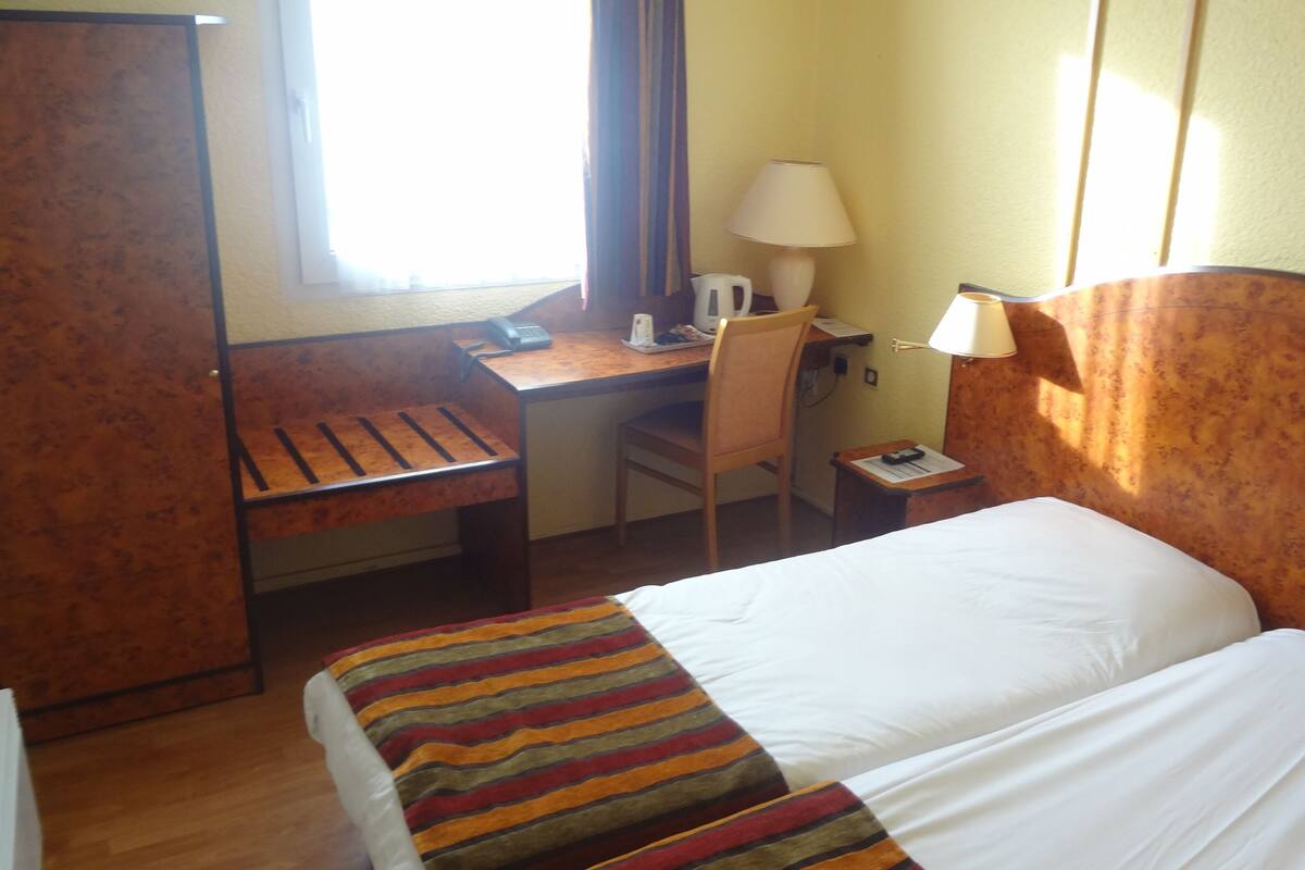 Quarto de hotel com uma cama, uma escrivaninha com cadeira, uma luminária e um telefone.