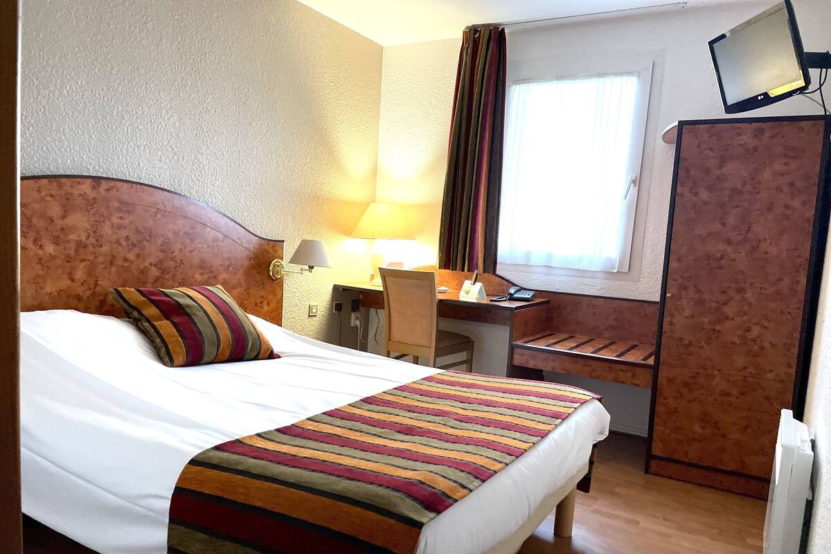Quarto de hotel com cama, escrivaninha, cadeira, abajur, televisão e janela com cortinas.